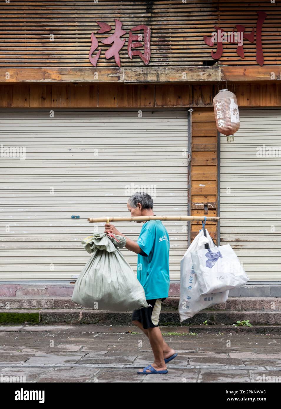 A Chinese man carries on his shoulders heavy sacks attached to a bamboo ...