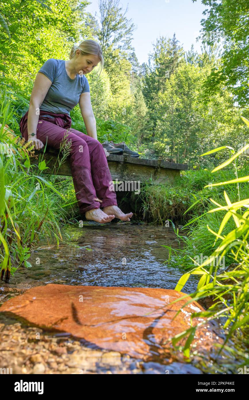 Woman sitting by stream holding feet in water on hiking trail ...