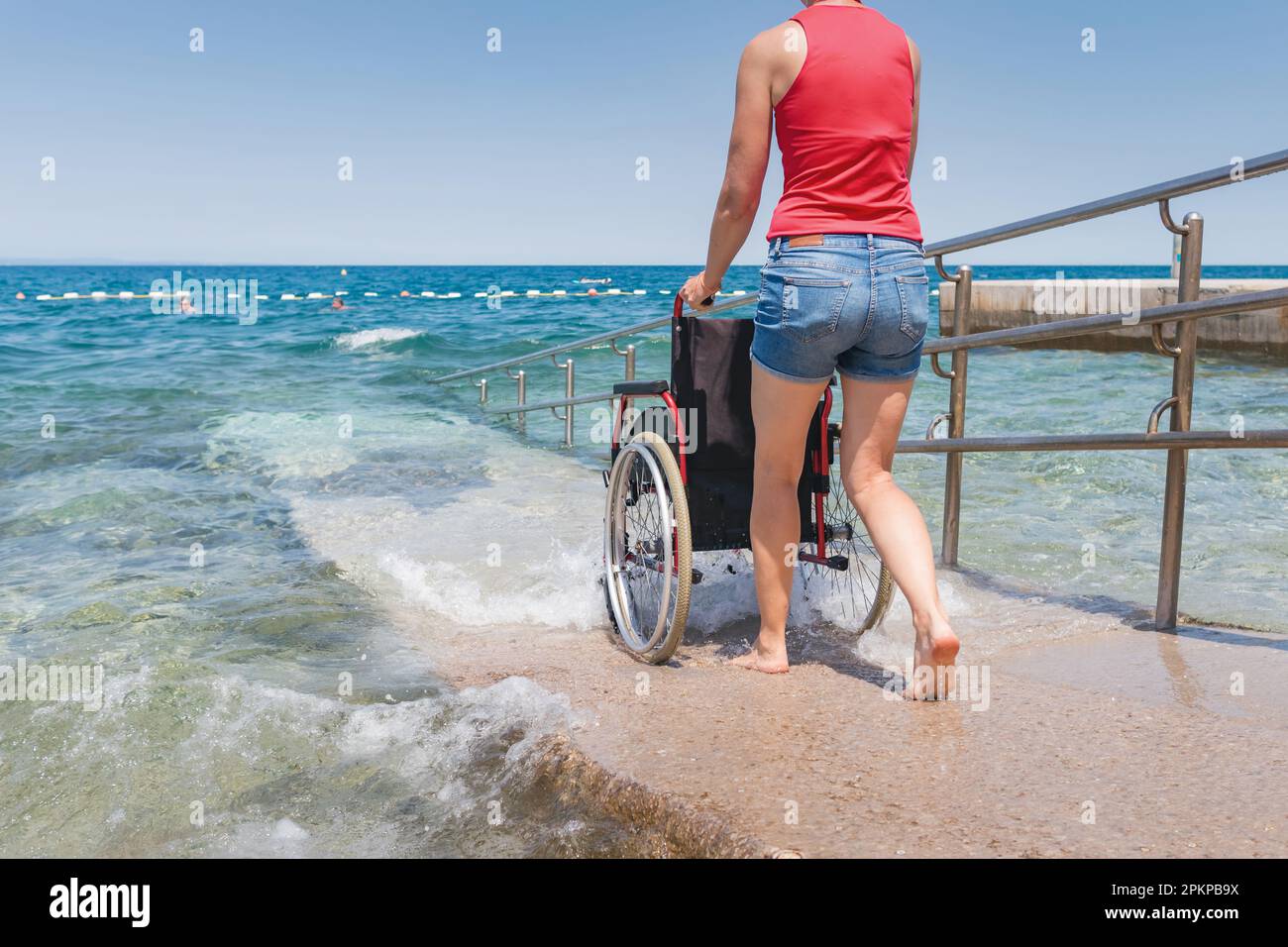 Woman assistant pushing wheelchair at sea on accessible beach with ramp ...
