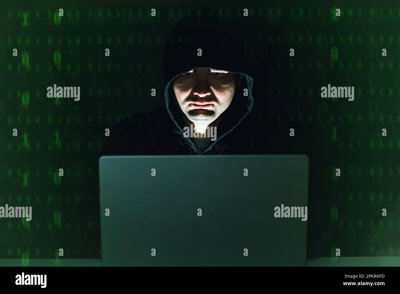 Hacker in the hoodie sitting in the front of his laptop at night, green ...