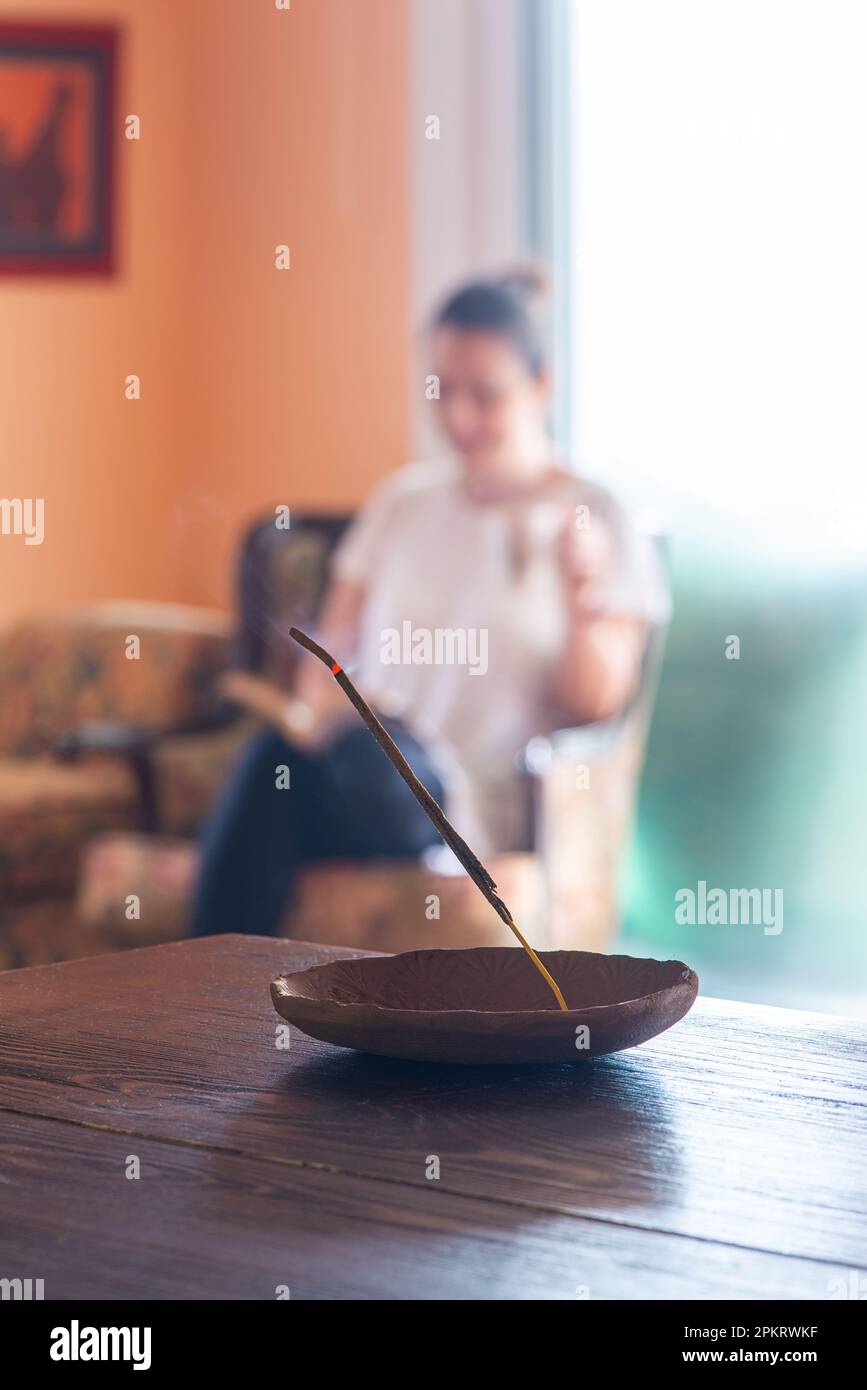 Incense stick burning with a woman sitting crosslegged in the background in a cozy room with ...