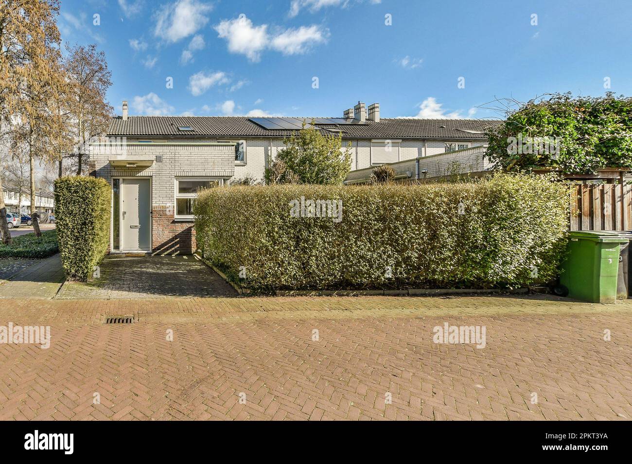 a house with some bushes in the front yard and a green trash can on the ...