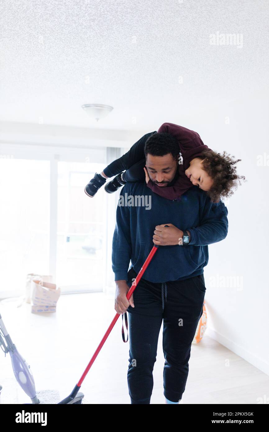 Black father cleaning the house, sweeping, and toddler on shoulders ...