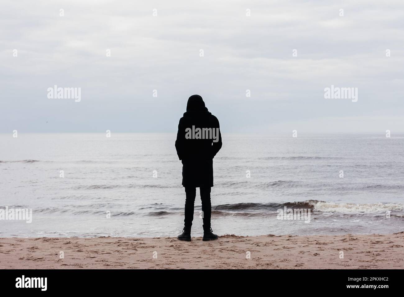 depressed man in black clothes with a hood stands by the sea in the ...