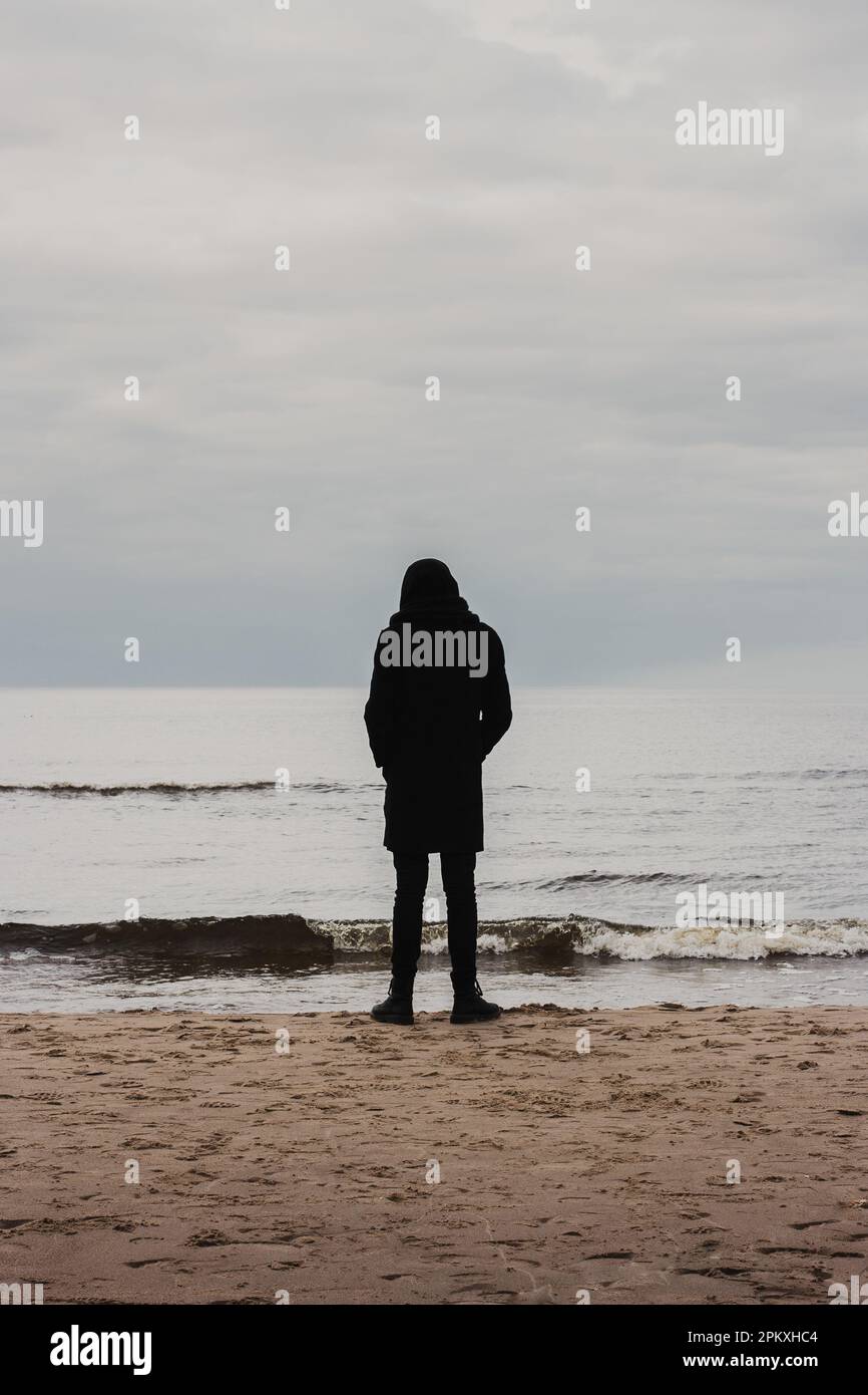 depressed man in black clothes with a hood stands by the sea in the ...