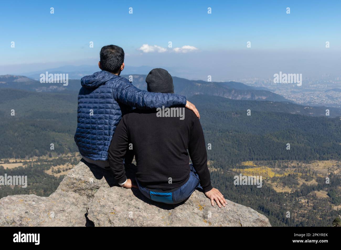 Back view of anonymous male travelers sitting on edge of Pico del Aguila mountain and hugging ...