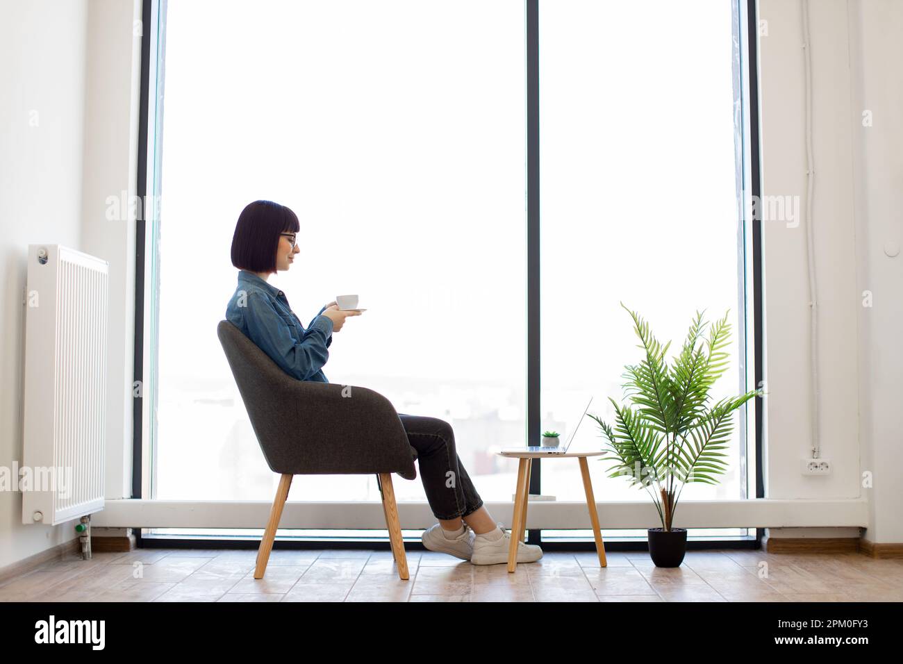 Side view of charming young woman sitting in armchair near table with ...