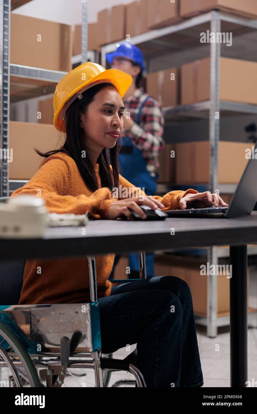 Warehouse employee in wheelchair checking product management system on laptop. Shipment manager ...