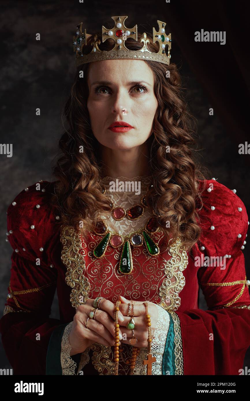 medieval queen in red dress with rosary and crown praying on dark gray ...