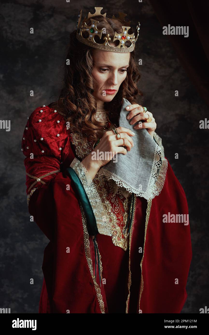 sad medieval queen in red dress with handkerchief and crown on dark ...