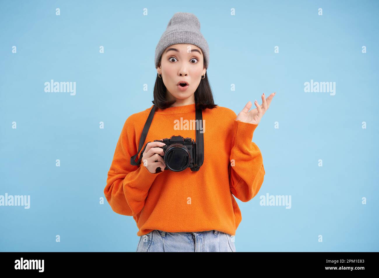 Surprised asian girl in hat, holds digital camera, looks amazed and ...