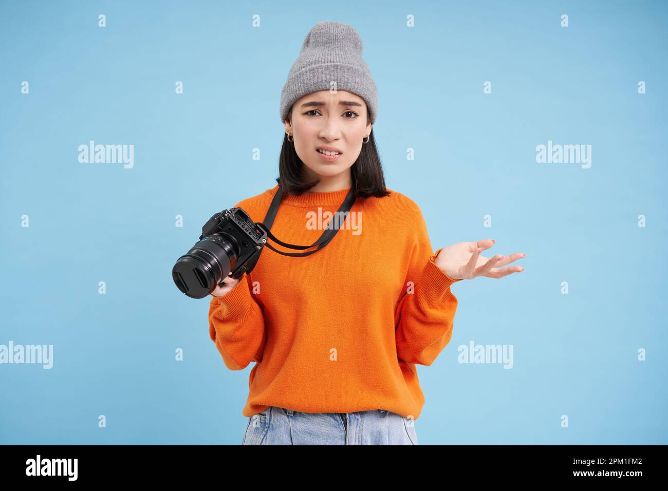 Young confused asian girl in hat, holds digital camera and shrugging ...