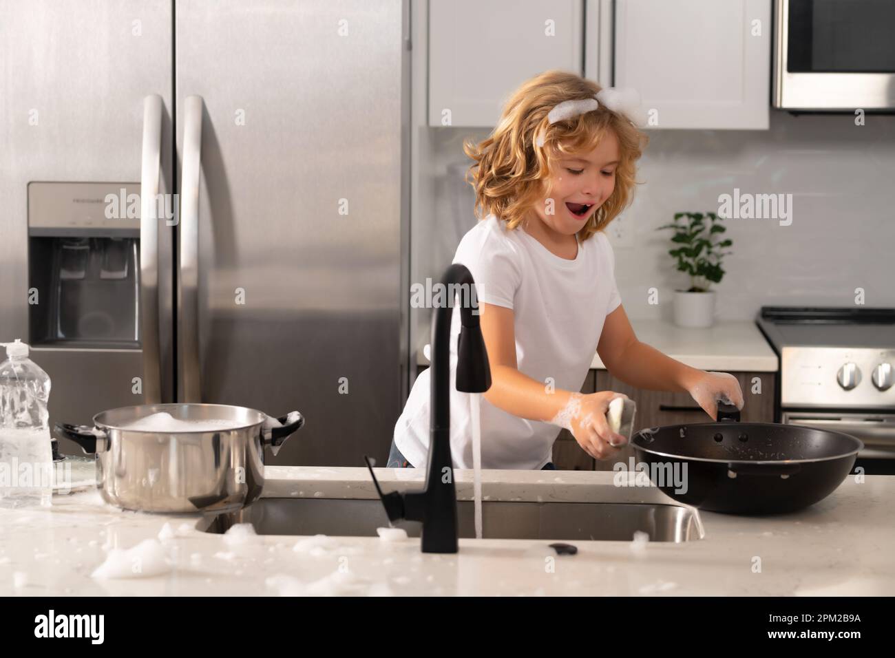 Excited child boy washing the dishes in the kitchen sink. Child with sponge with dish washing ...