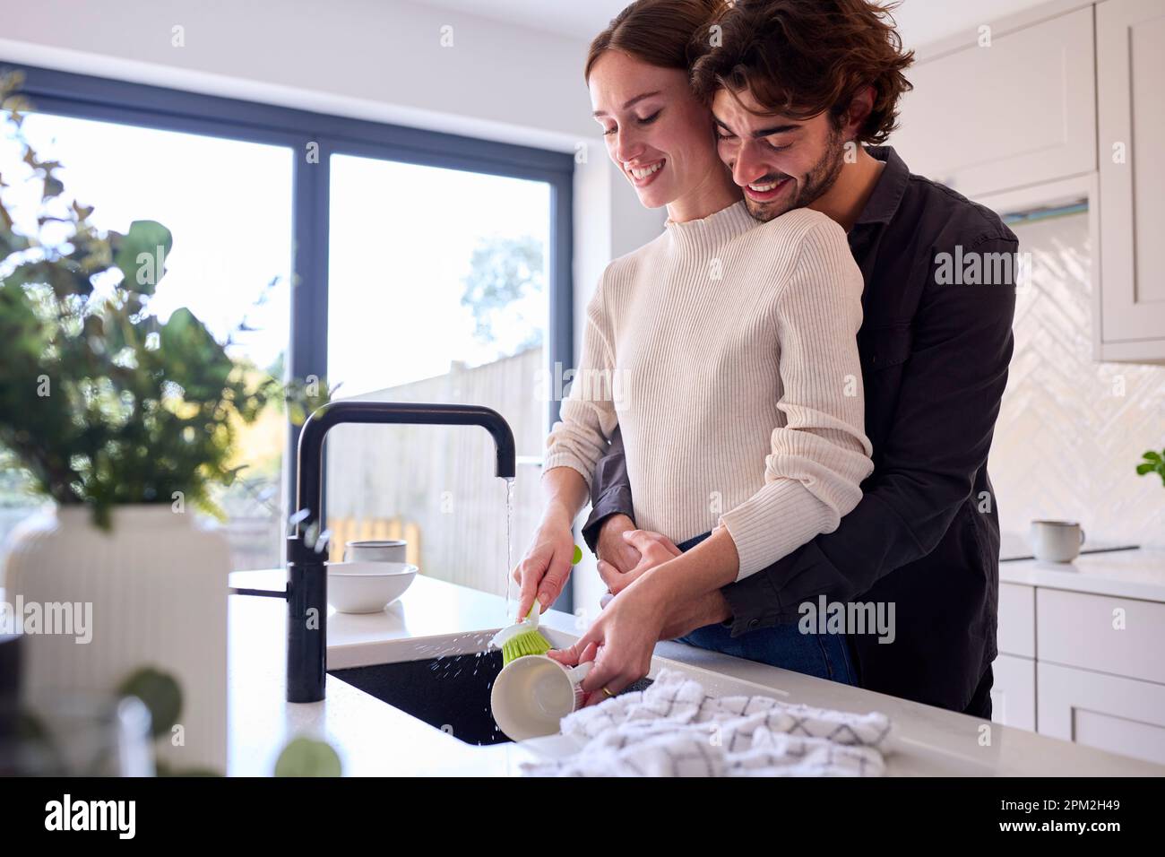 Couple Hugging At Home In Kitchen As Woman Does Washing Up Stock Photo ...