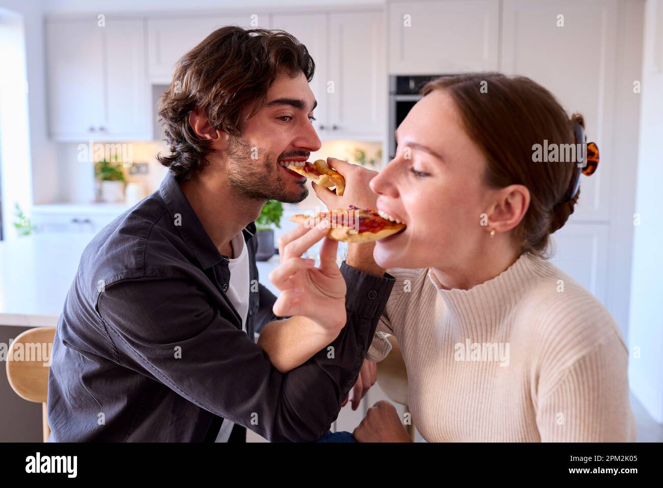 Couple In Kitchen At Home Linking Arms To Eat Homemade Pizza Sitting At ...