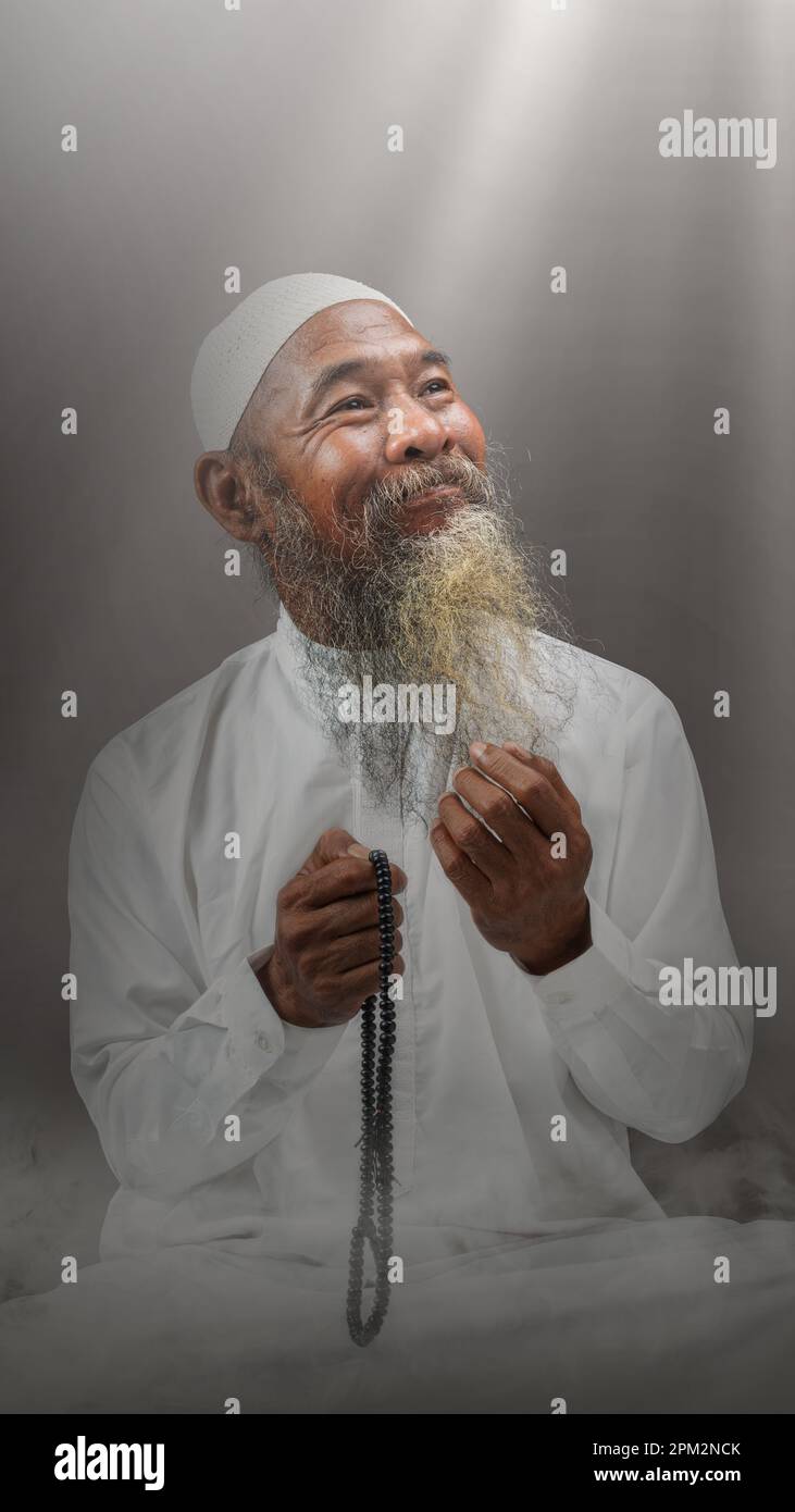 Muslim man with a beard wearing a white cap praying with prayer beads ...