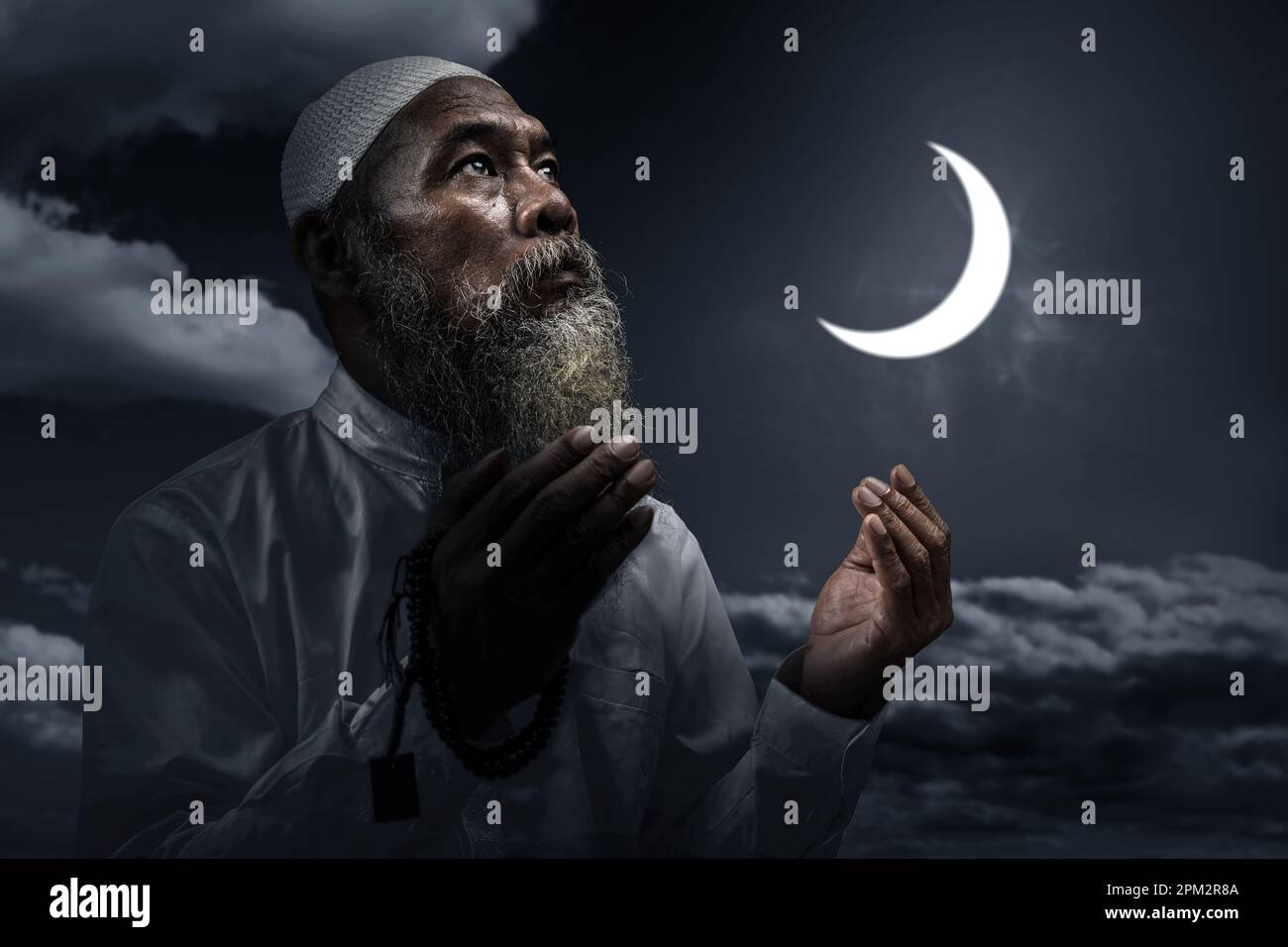 Muslim man with a beard wearing a white cap praying with prayer beads ...