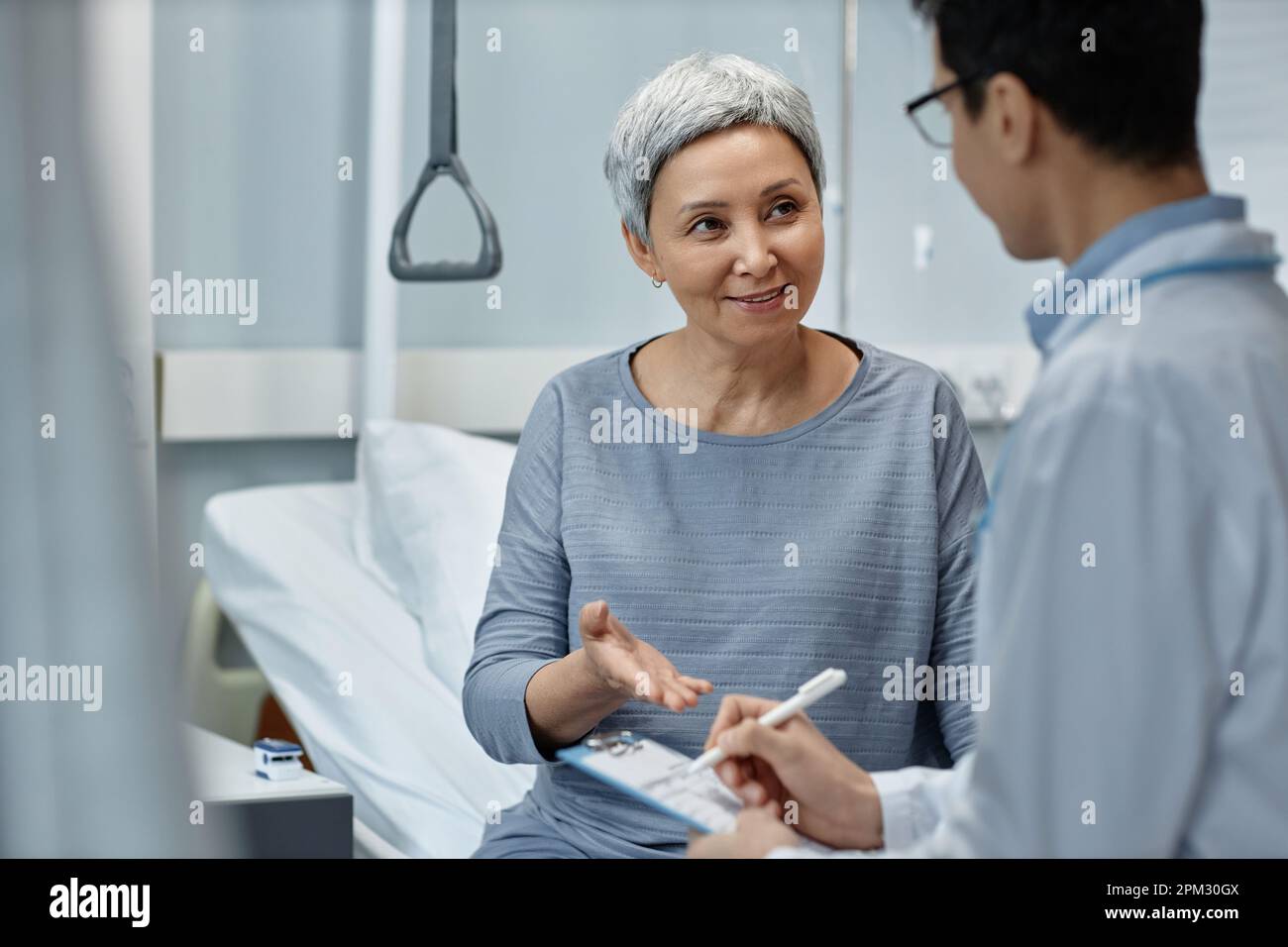 Young doctor asking questions to senior woman while they sitting on bed ...