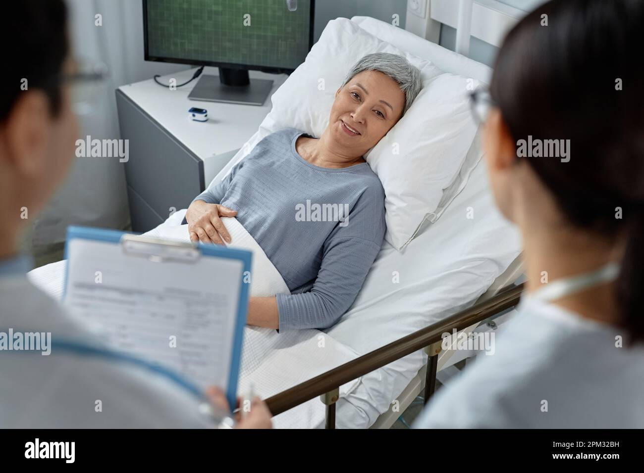 Mature woman lying on bed in ward and discussing her treatment together with doctor and nurse ...
