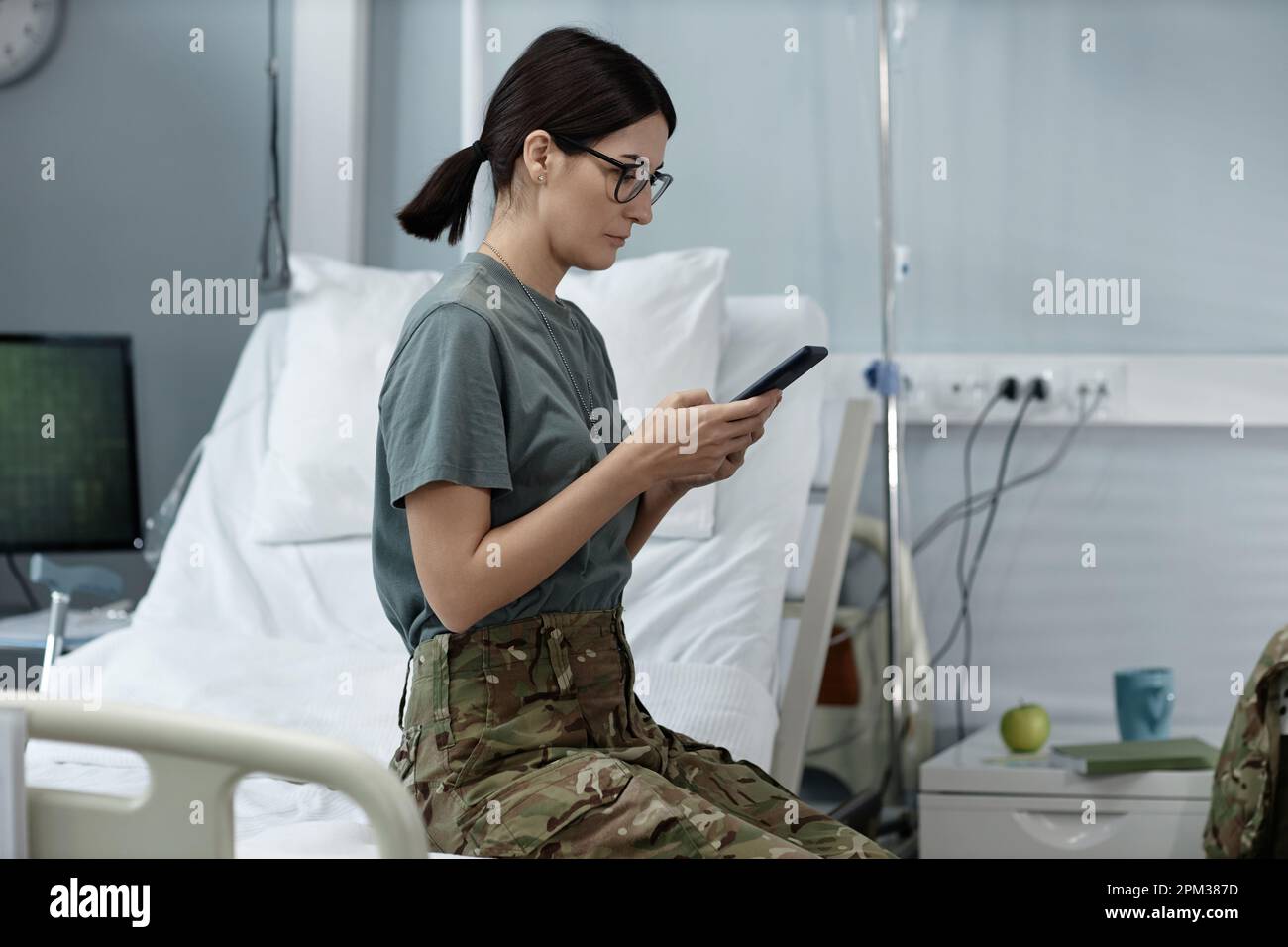 Young female soldier typing message on smartphone while sitting on bed ...