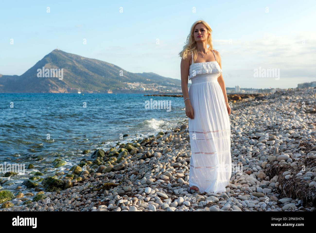 pretty woman in white dress, walking along a pebble beach, at sunset ...