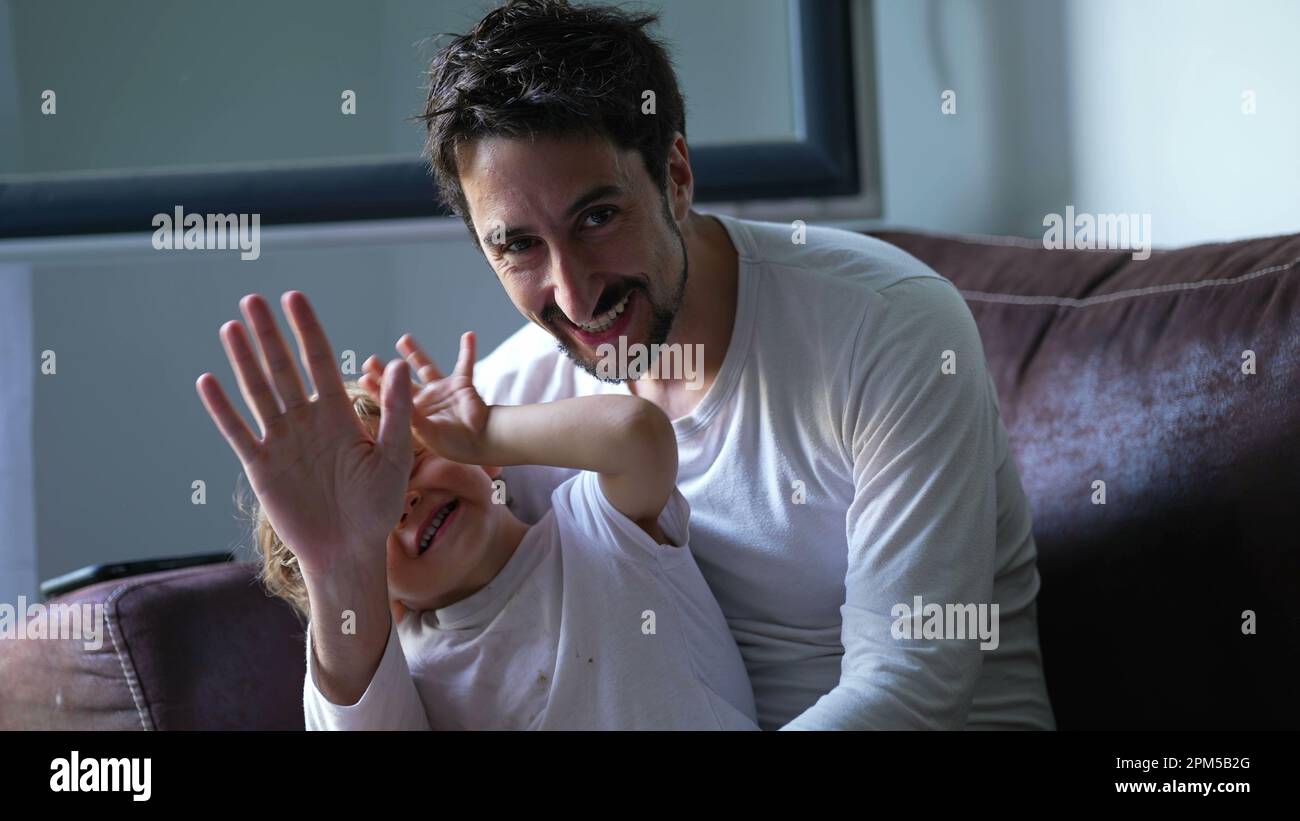 Father and child waving hello to camera sitting on sofa indoors. Dad ...