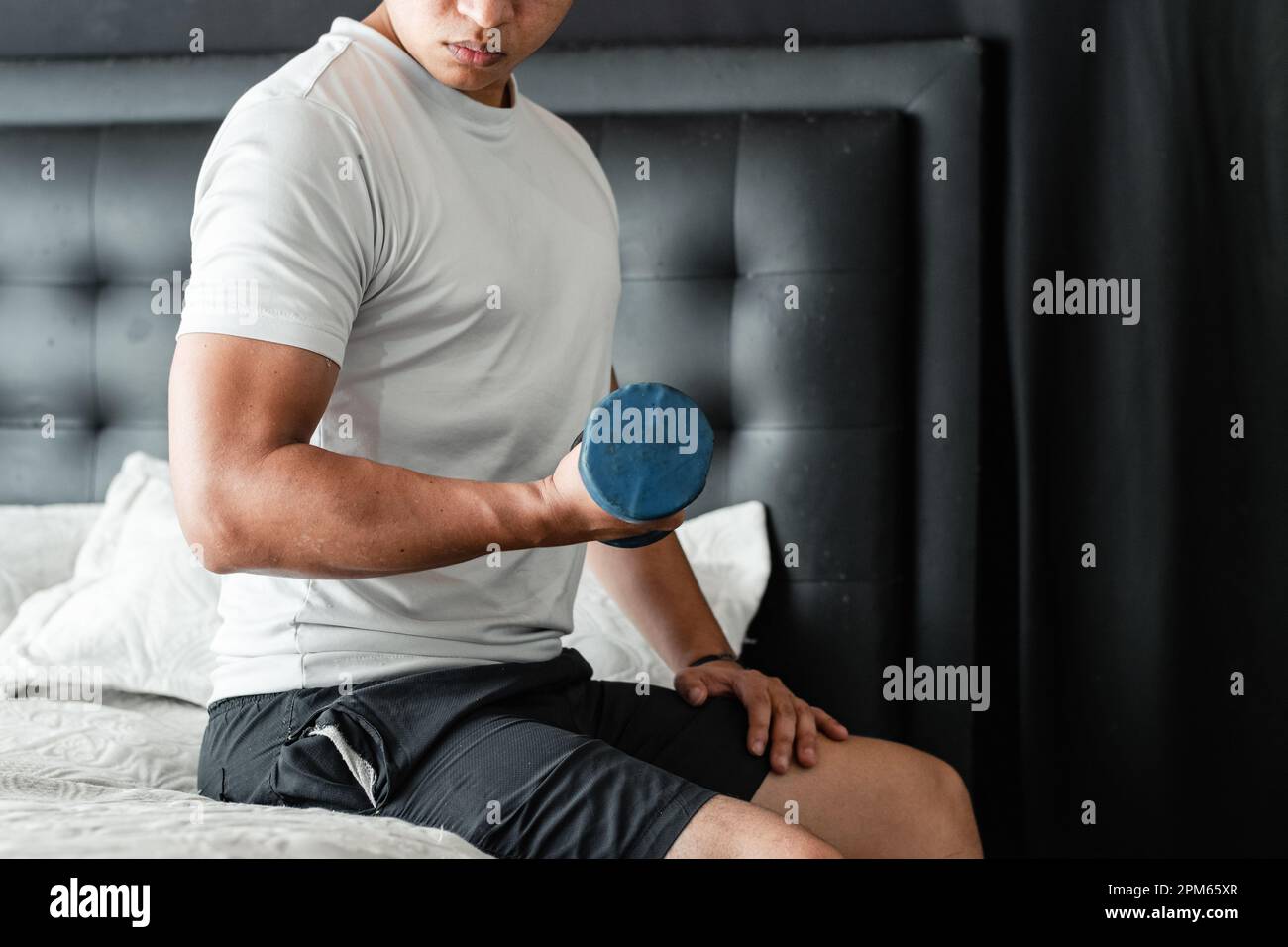 Young Latin Man Exercising Sitting On His Bed With A Blue Dumbbell