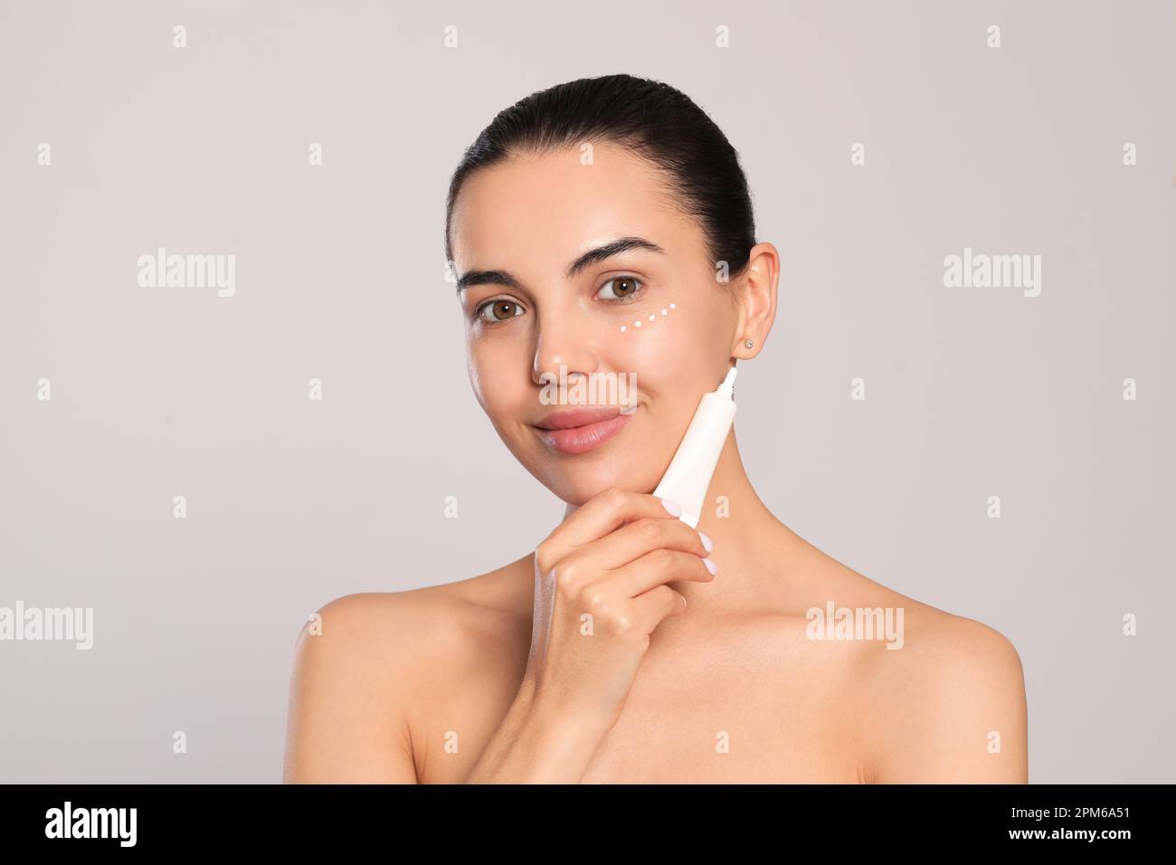 Beautiful young woman with cream on skin under eye against light grey ...