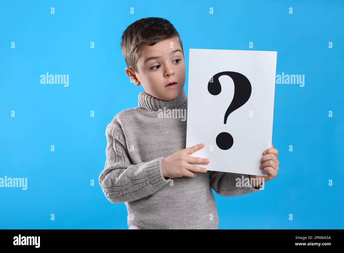 Emotional little boy holding paper with question mark on light blue ...