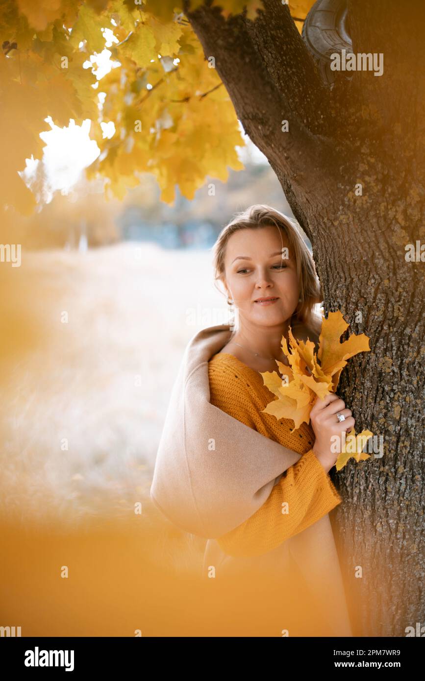 The blonde stands near the autumn tree. Thoughtful woman looks ahead ...