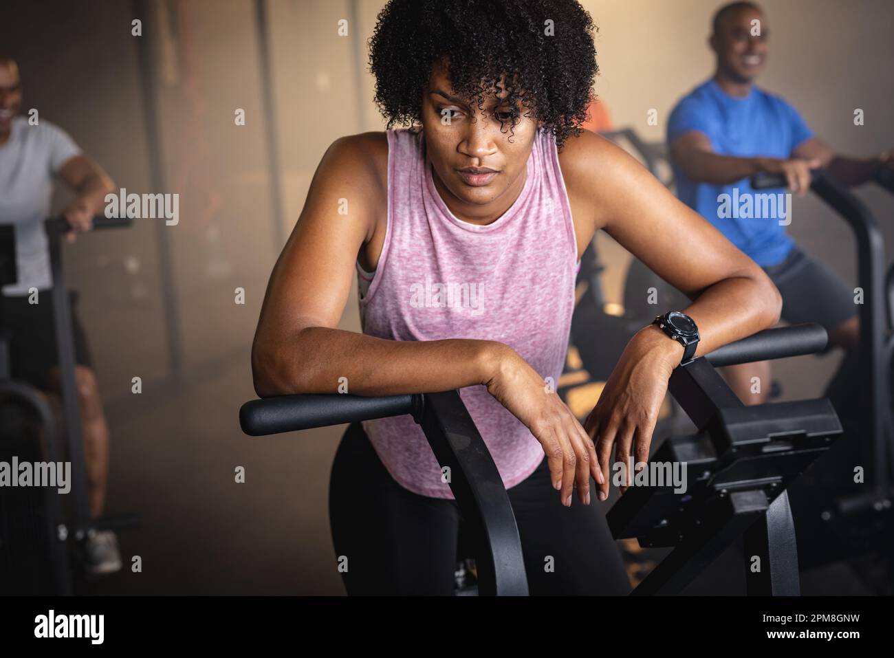 Tired biracial young woman with afro hair exercising on bike in gym ...