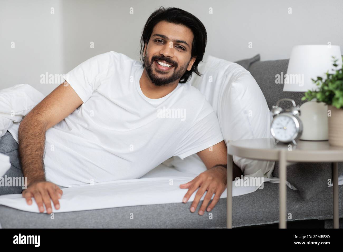 Cheerful young middle eastern male in white t-shirt lies on bed, enjoy ...