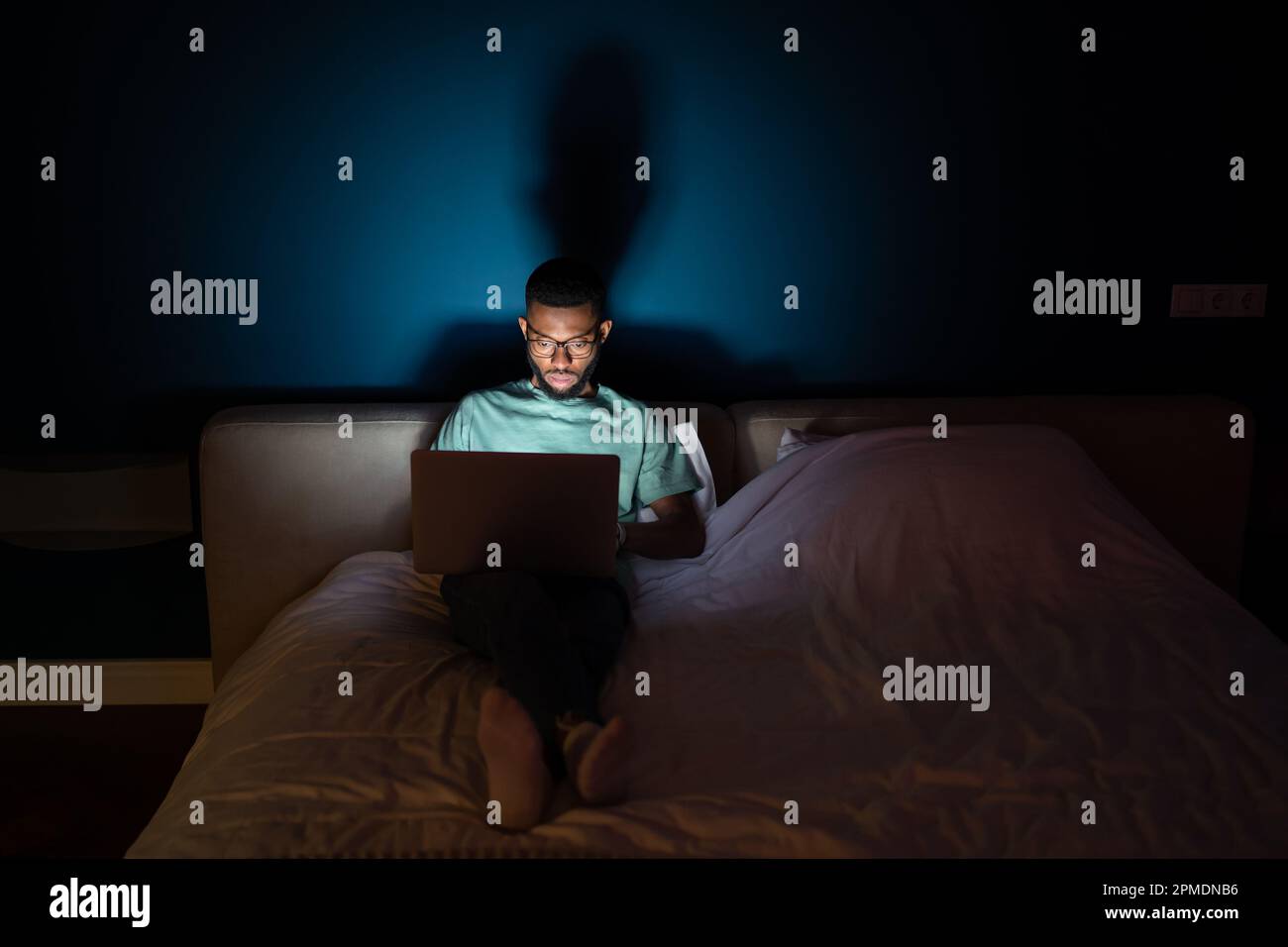 Young black man works on laptop lying in bed at night in dark looks ...