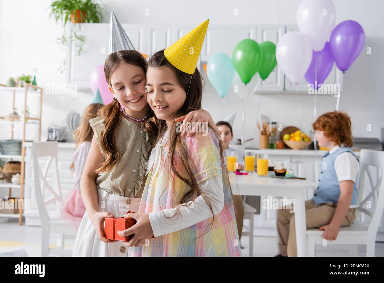 cheerful birthday girl holding present and hugging friend during ...
