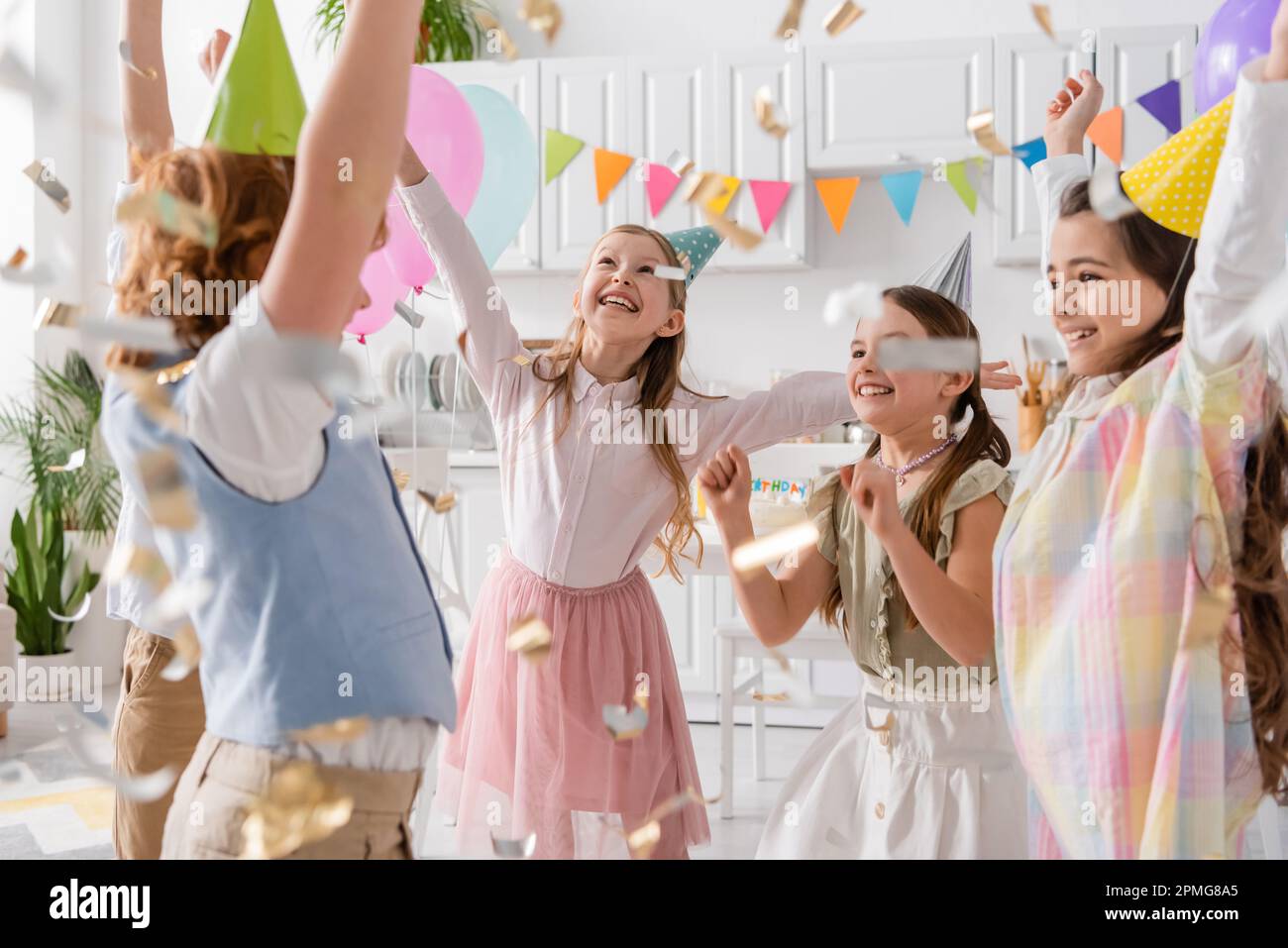 group of excited kids in party caps dancing under falling confetti ...