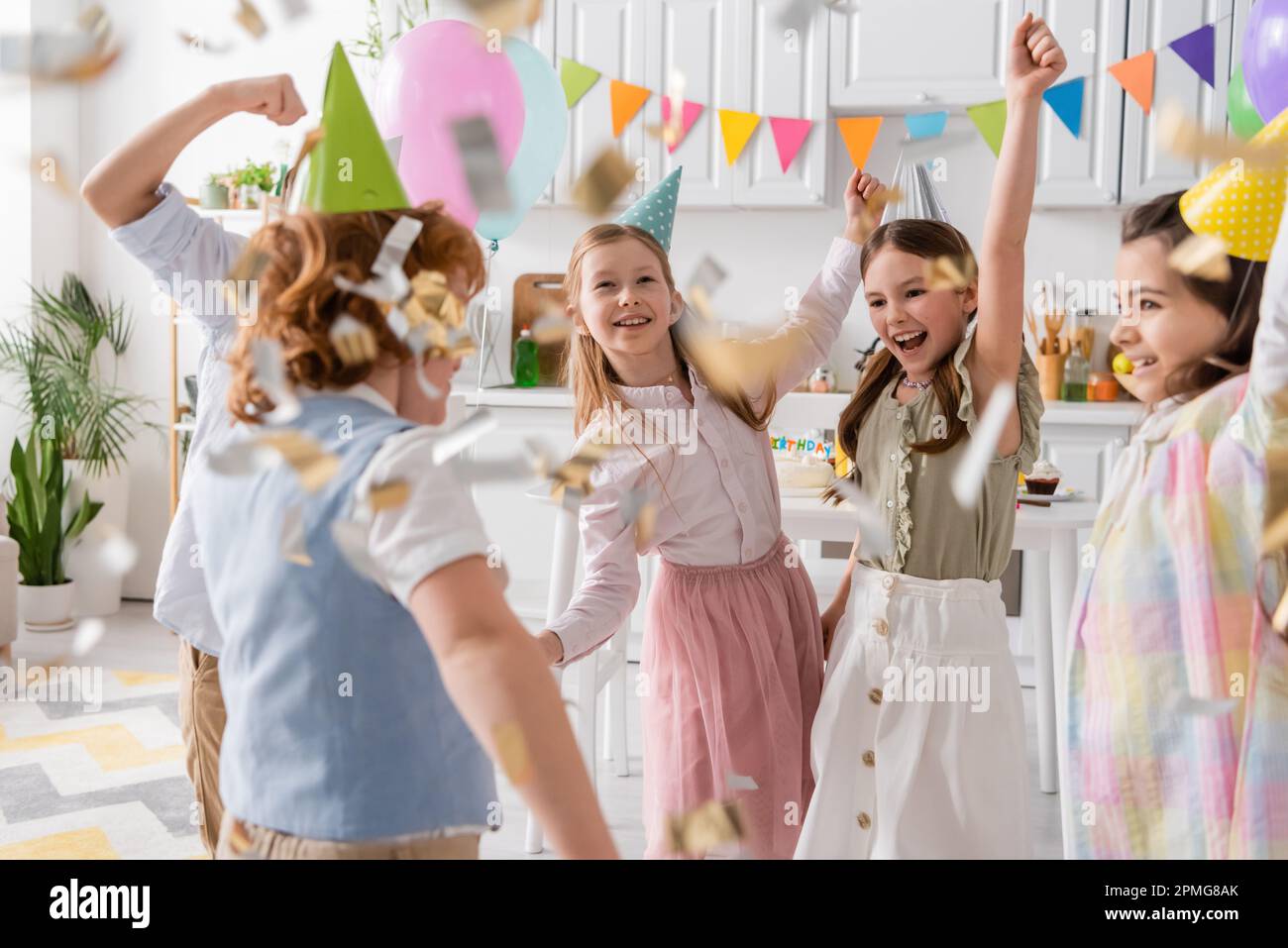 group of happy kids in party caps dancing under falling confetti during ...