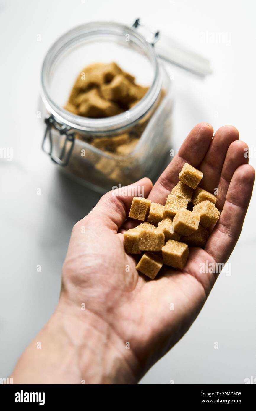 Brown sugar cubes in a man's hand and in the jar on white marble ...