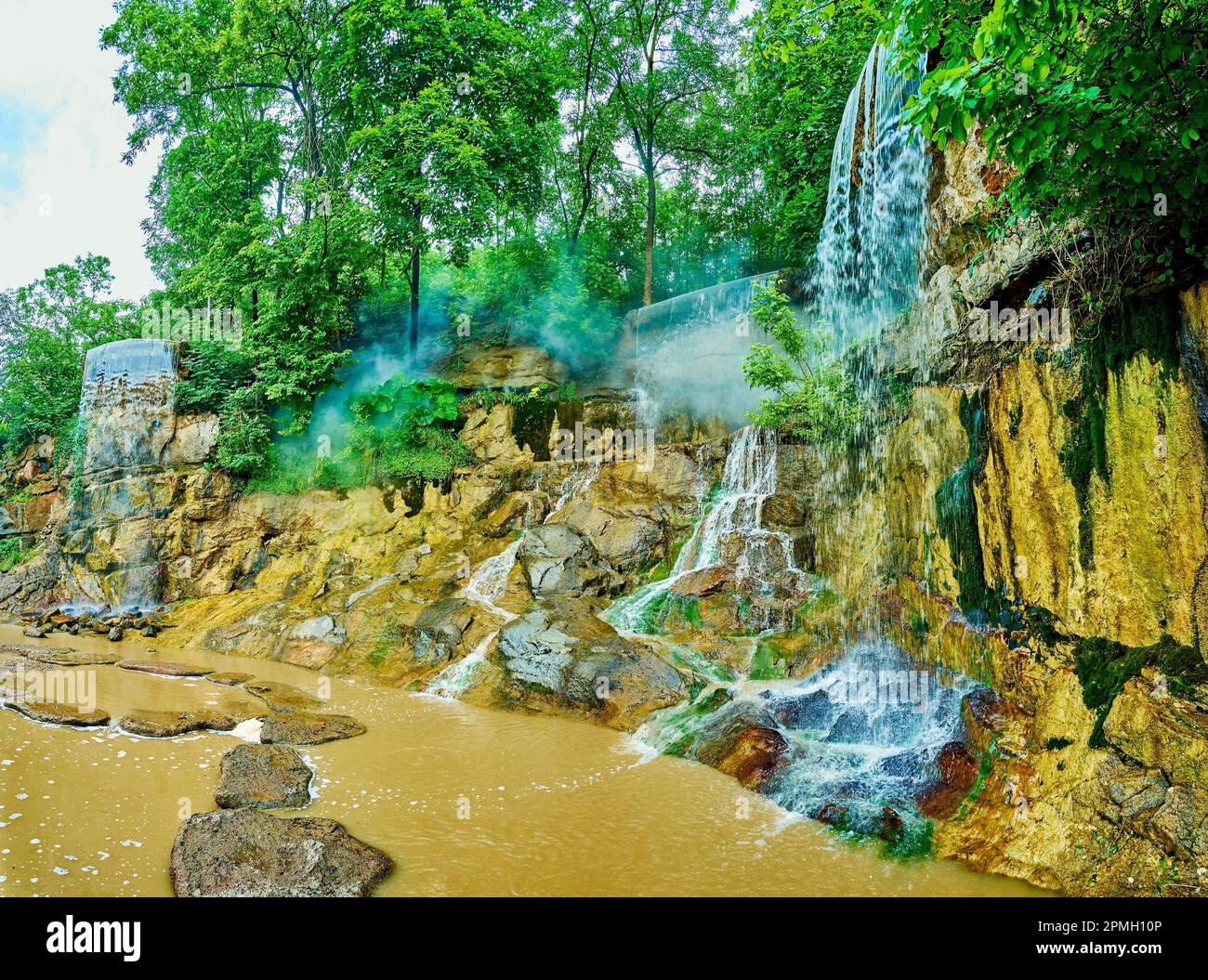 The waterfall on the rocky cliff edge among the lush greenery of ...