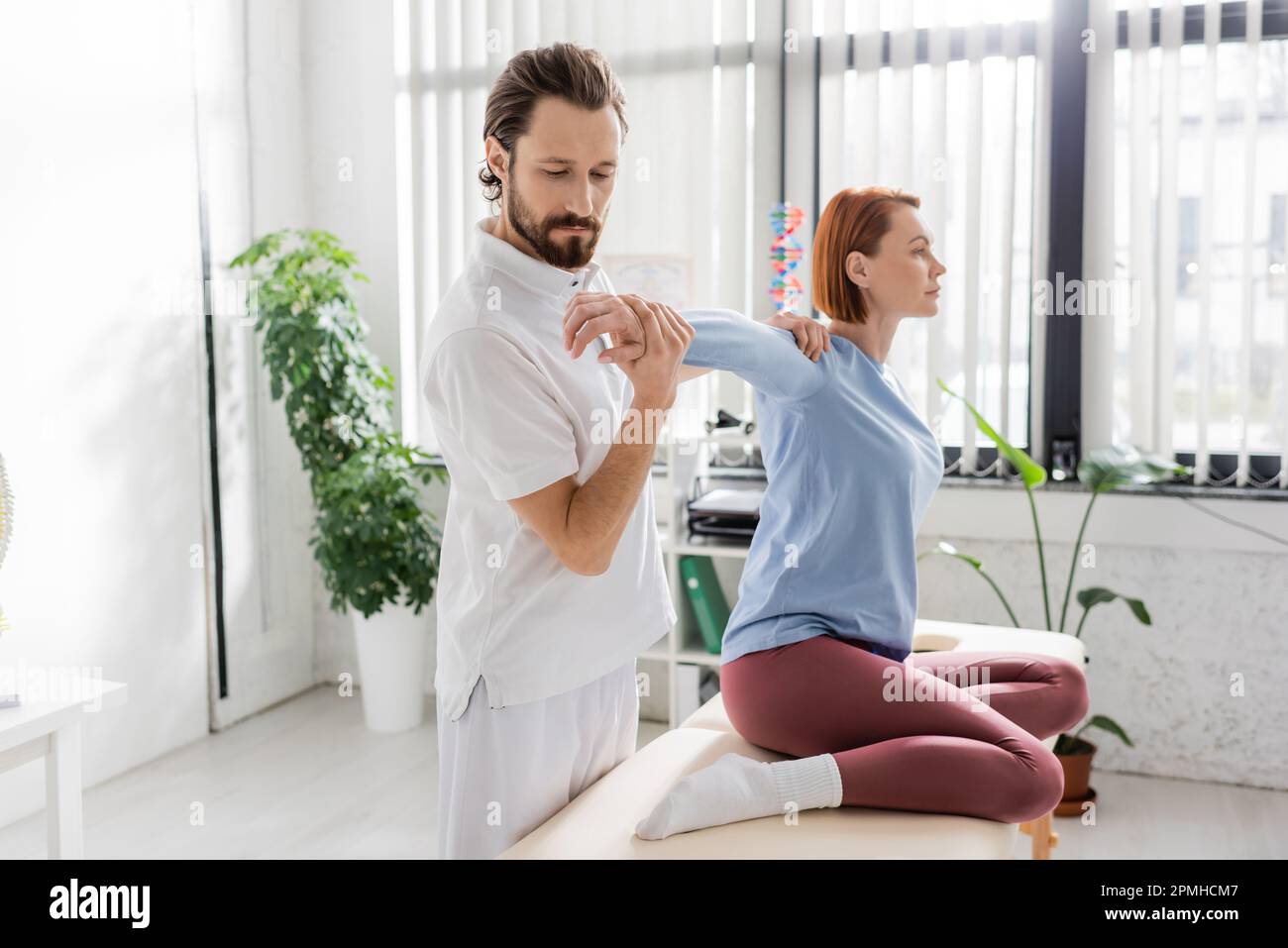 bearded osteopath stretching arm of woman sitting on massage table in ...