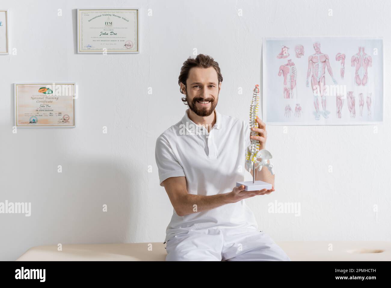 joyful chiropractor smiling at camera while sitting with spine model in ...