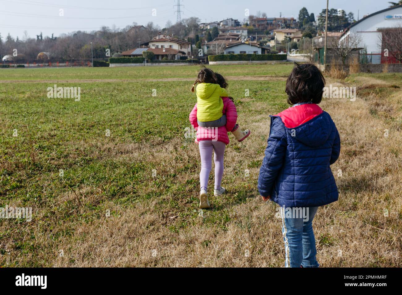 rear view of three children walking home with small child riding ...