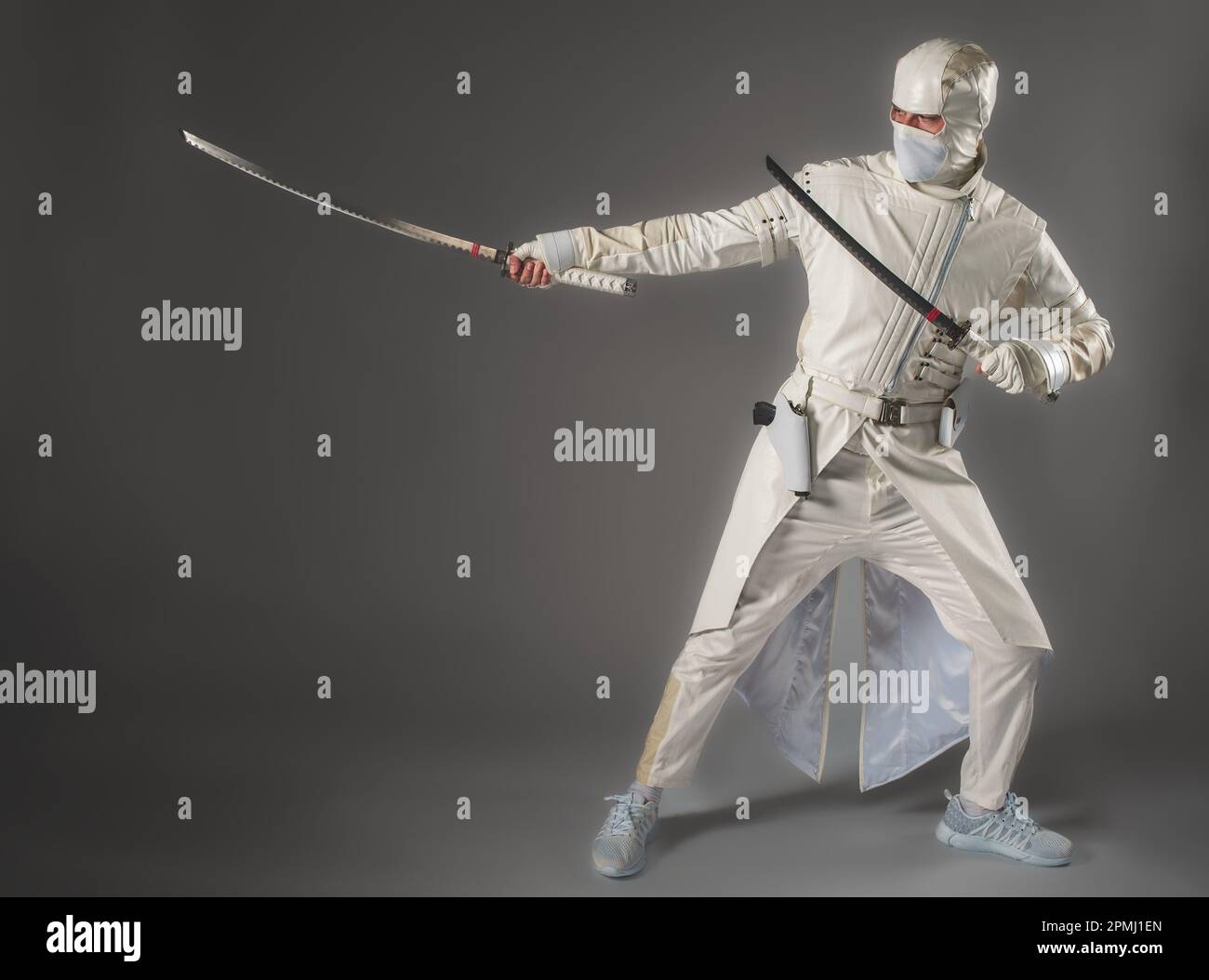 A man in the form of a combat ninja poses with two swords on a gray ...