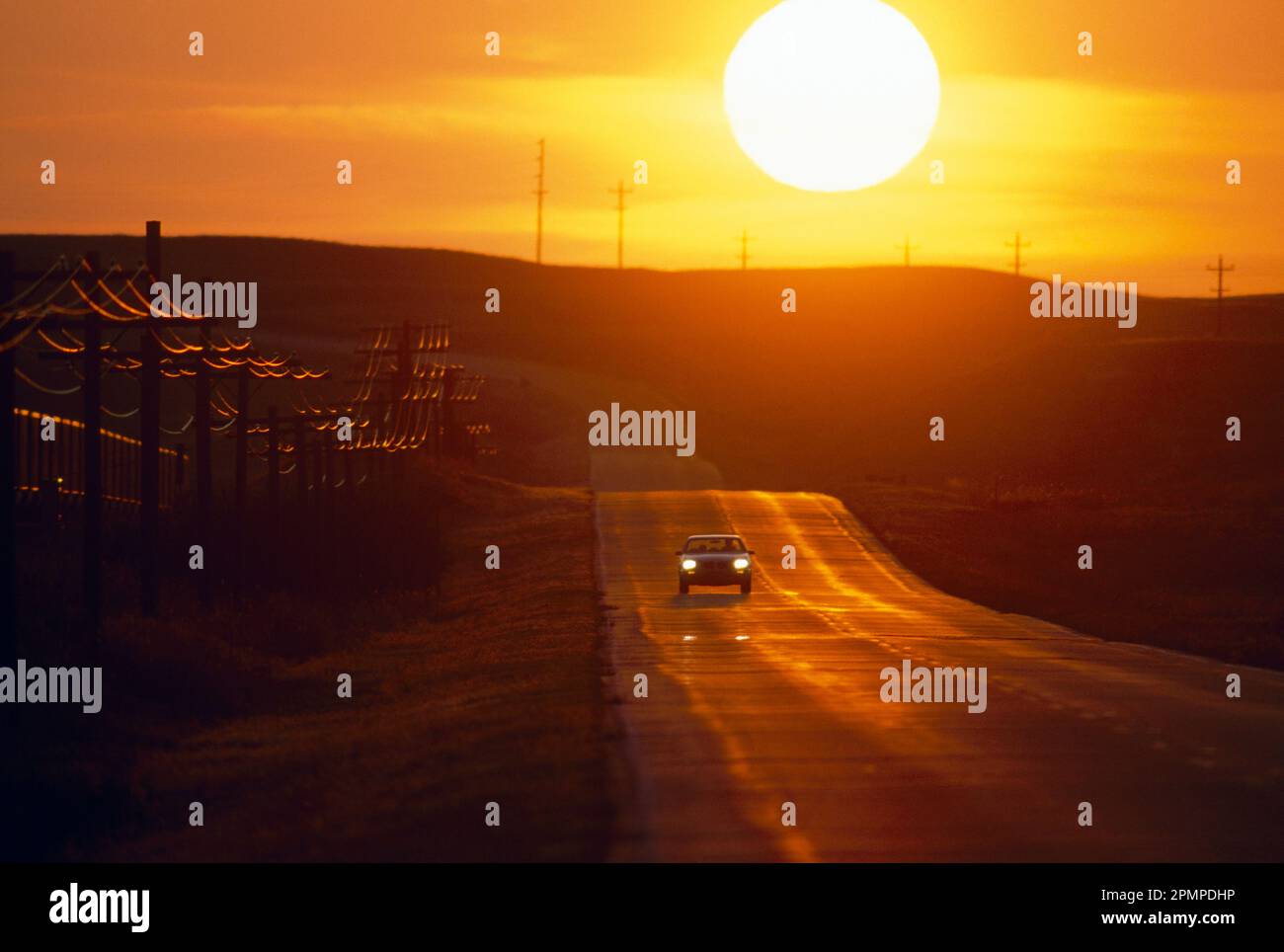 Lone car driving along a rural highway below a large, orange sun ...