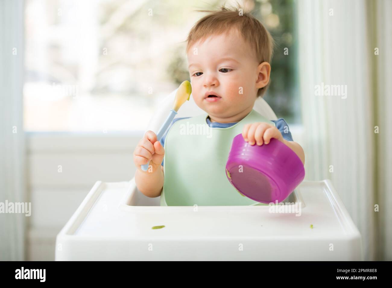 Cute baby eating first solid food, infant sitting in high chair. Child tasting vegetables at the ...