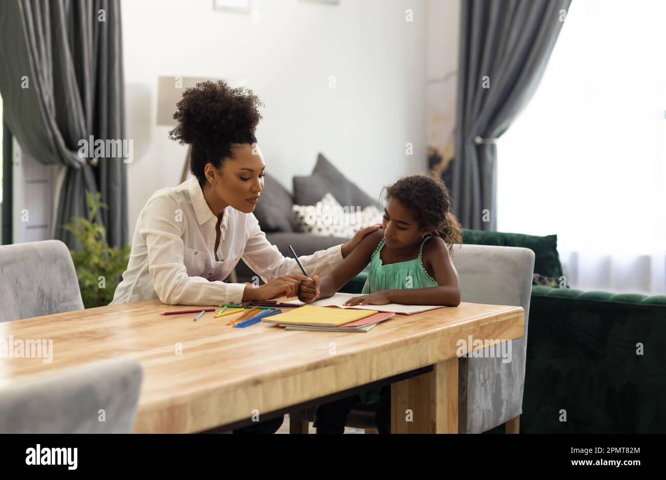 Focused biracial mother sitting at dining table helping her daughter ...