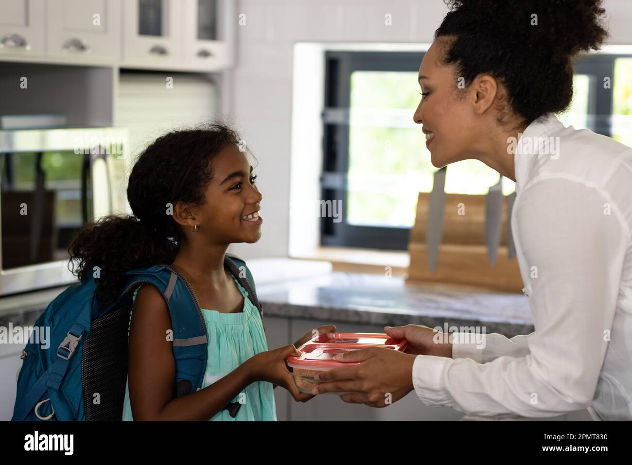 Happy biracial mother giving school lunchbox to her smiling daughter ...