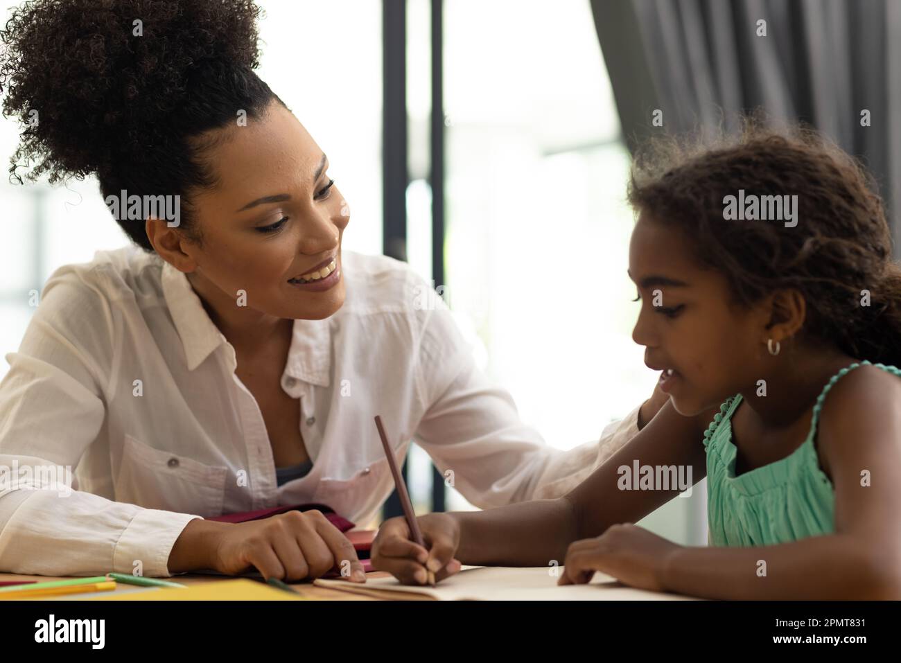 Smiling biracial mother sitting at dining table helping her daughter ...