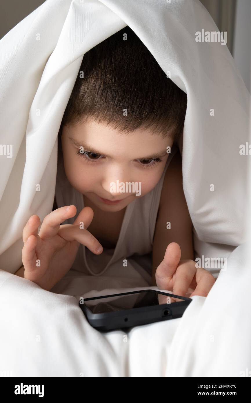 A 5 year old boy plays games on his phone while lying in bed. Gadgets ...