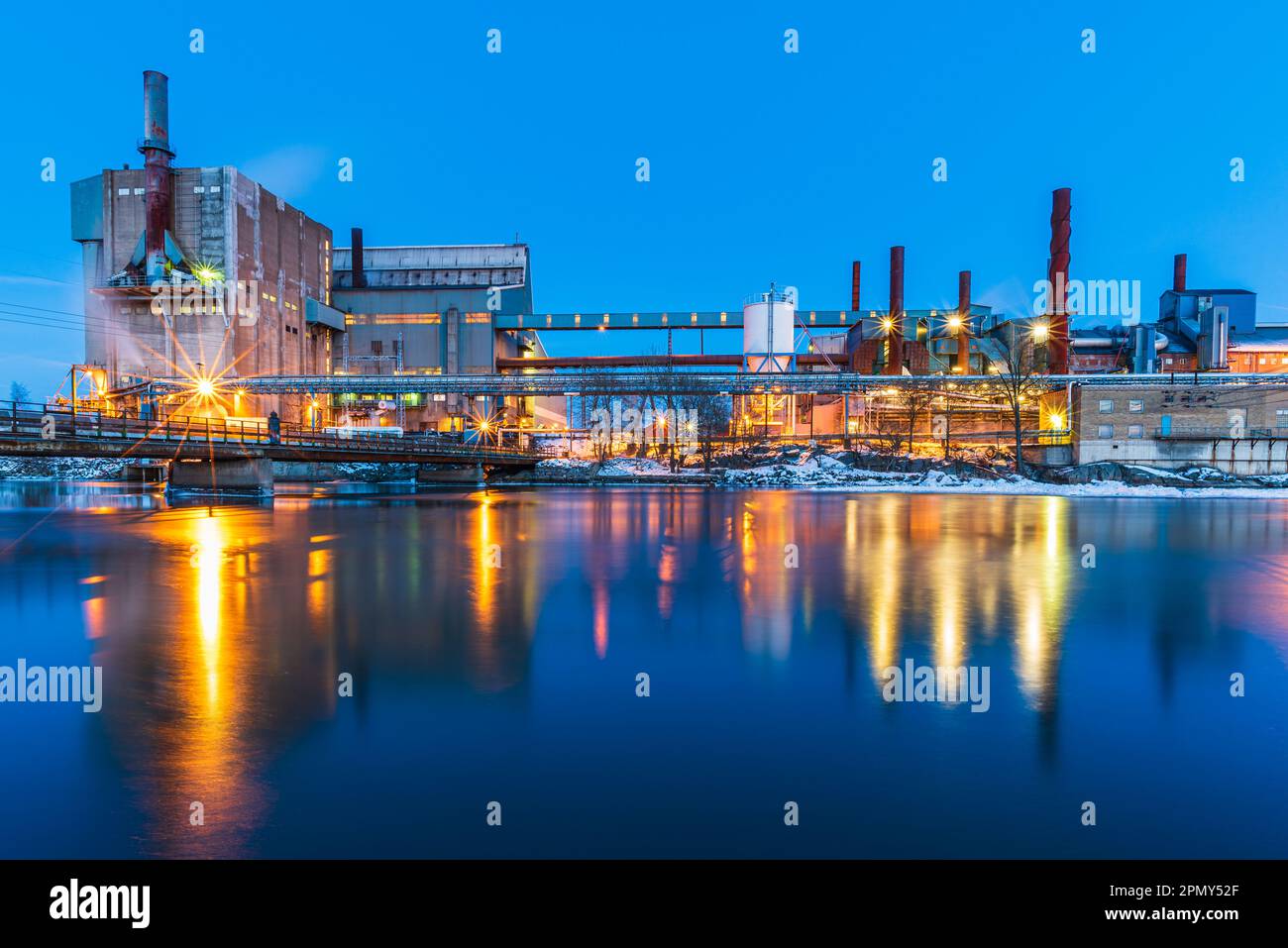 At dusk, the illuminated factory casts a reflection onto the river ...