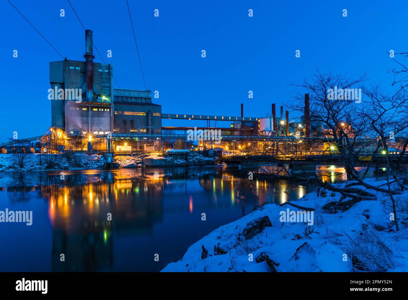 At dusk, the illuminated factory casts a reflection onto the river ...